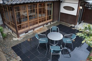 Um pátio com uma mesa redonda e quatro cadeiras, uma janela de madeira com padrão quadriculado e uma pequena área de jardim com pedriscos e plantas.