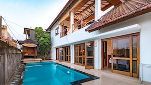 Uma área de piscina com piscina azul límpida, deck de madeira e um edifício com grandes portas de vidro e telhado em azulejos.