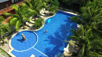 Uma piscina ampla e azul, com um trecho circular e uma estátua no centro, rodeada por palmeiras e um deque de madeira.
