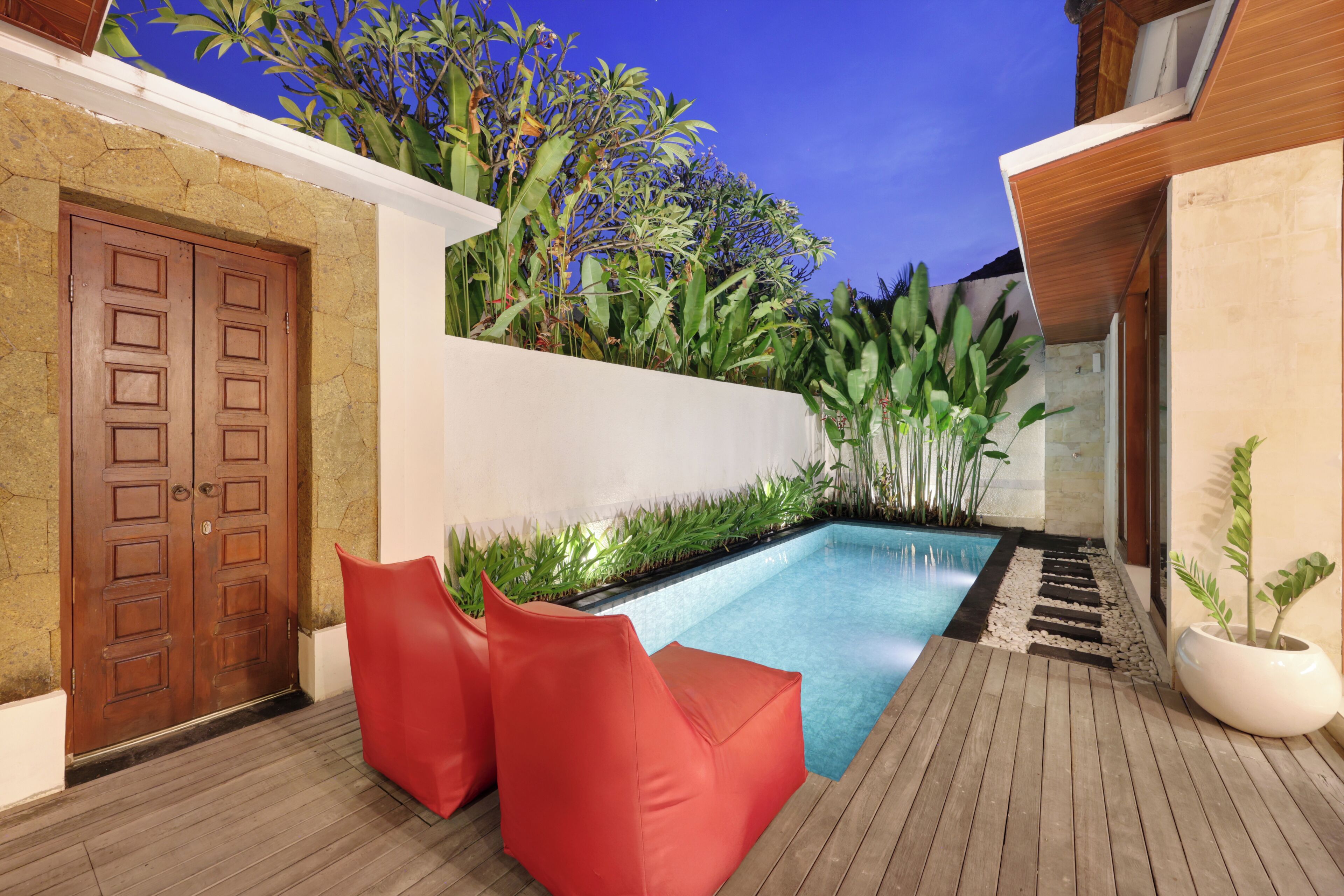Uma área de piscina com deck de madeira, cadeiras vermelhas e um muro de pedra com plantas.