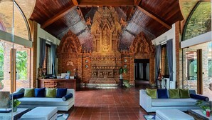 Uma sala de estar ampla, com um grande altar de madeira ricamente entalhado no centro, cercado por assentos confortáveis e piso ladrilhado.