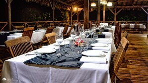 Restaurante com mesas postas para refeições, cadeiras de madeira e estrutura de telhado de palha.