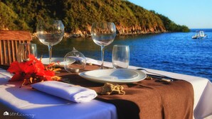 Uma mesa posta com taças de vinho, pratos e um guardanapo dobrado, com vista para um corpo d'água e um barco ao longe.