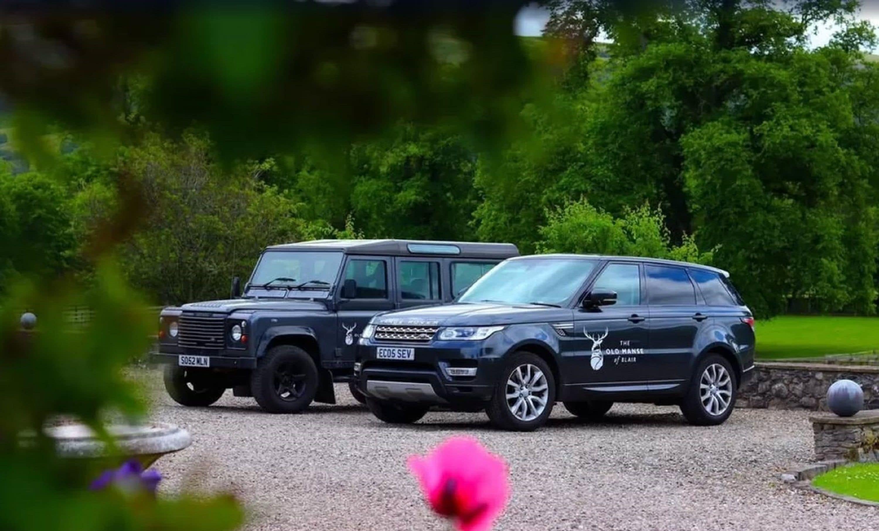 Dois SUVs estacionados em um caminho de cascalho, com um jardim e árvores ao fundo.