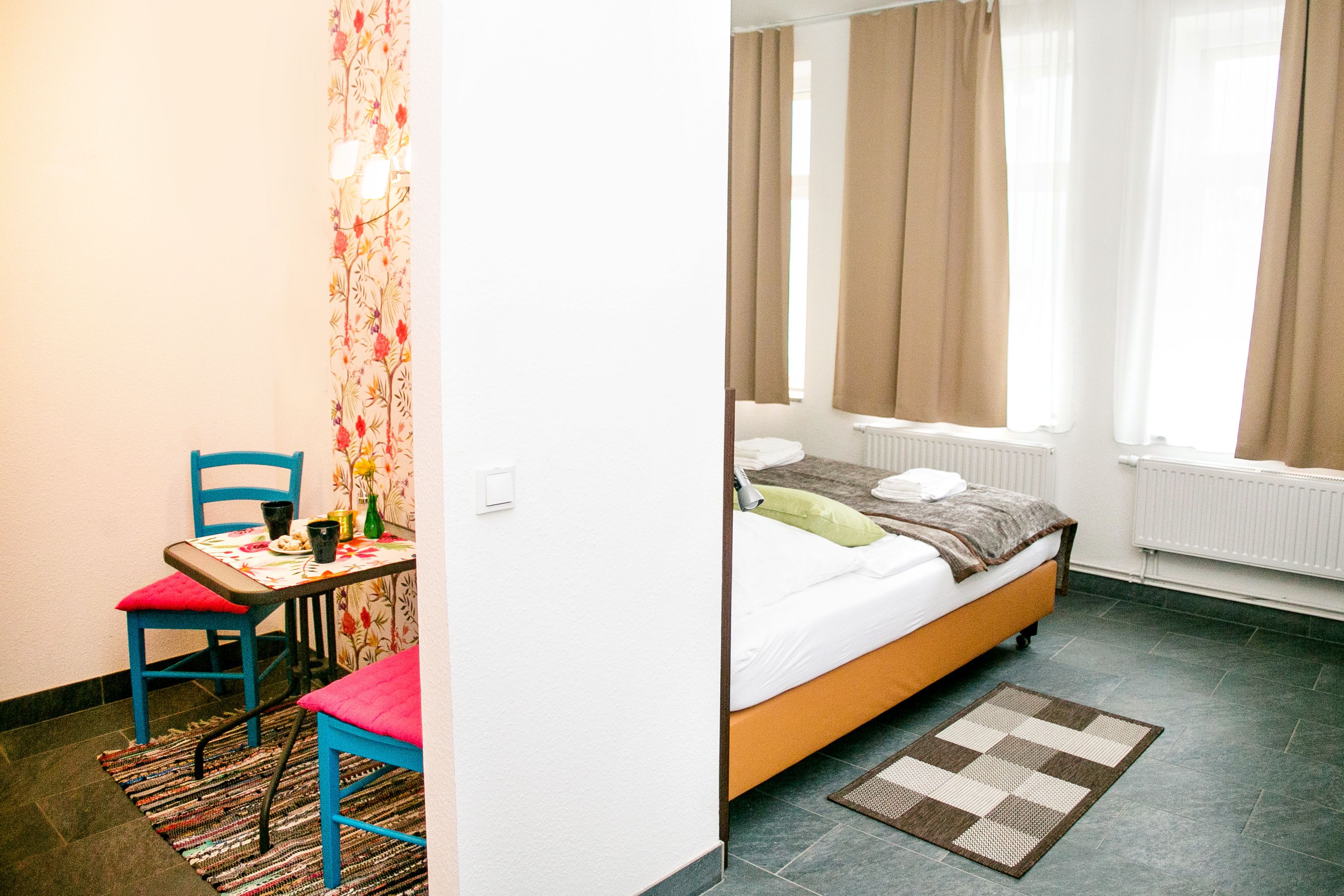 Quarto com cama, janela com cortinas, radiador e uma pequena mesa com cadeira.