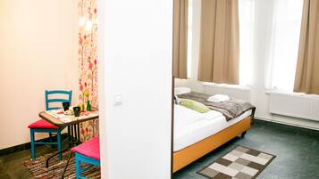 Quarto com cama, janela com cortinas, radiador e uma pequena mesa com cadeira.