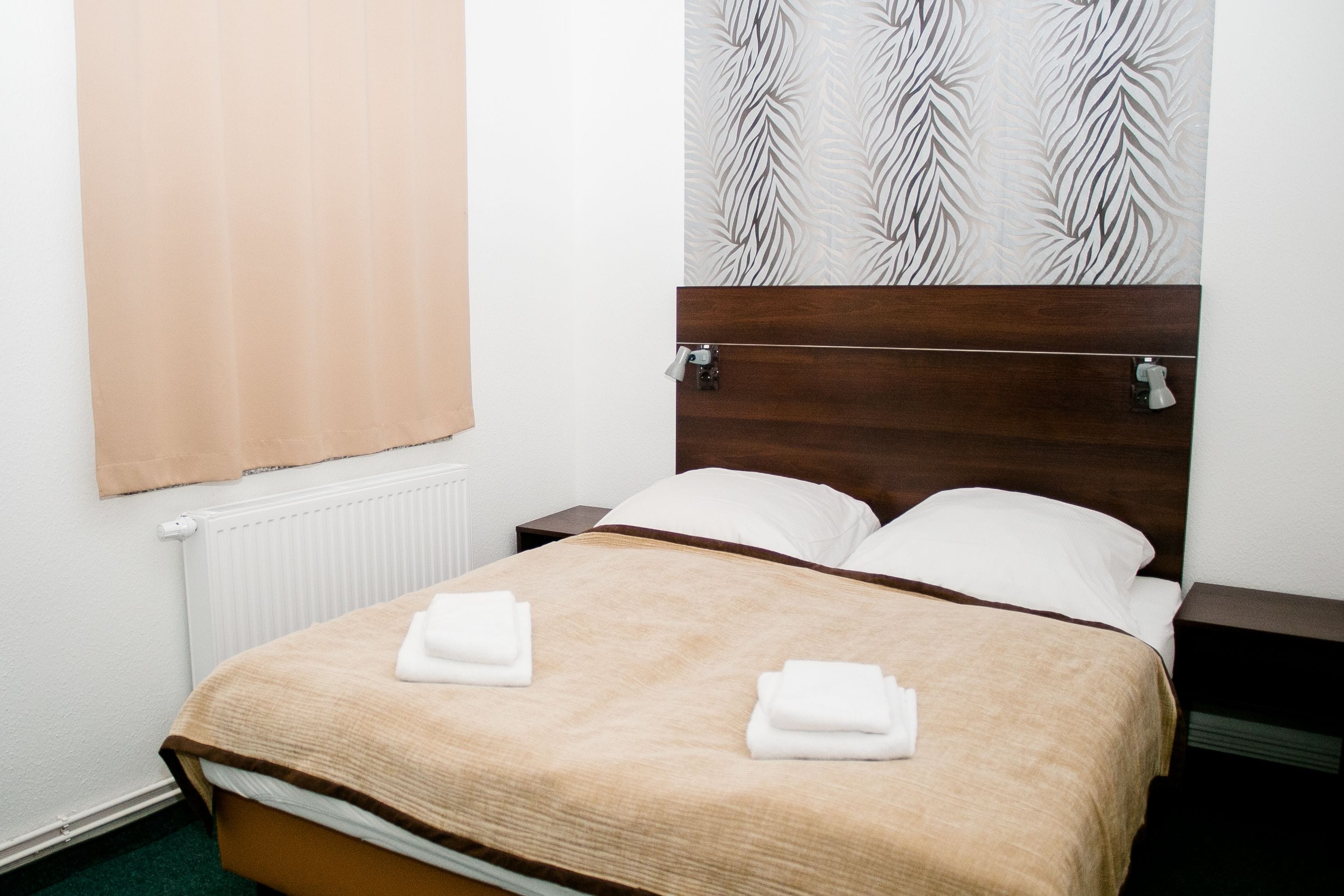 Quarto de hotel com cama, duas almofadas, cabeceira de madeira, mesa de cabeceira e radiador.