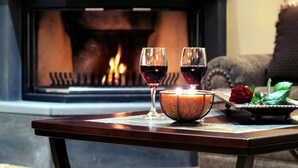 Um quarto aconchegante com lareira, dois copos de vinho tinto, uma vela e uma rosa em uma mesa.