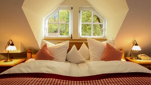Quarto com teto inclinado, duas janelas com molduras brancas, cama com roupa de cama branca e almofadas vermelhas xadrez, e luminárias de cabeceira.