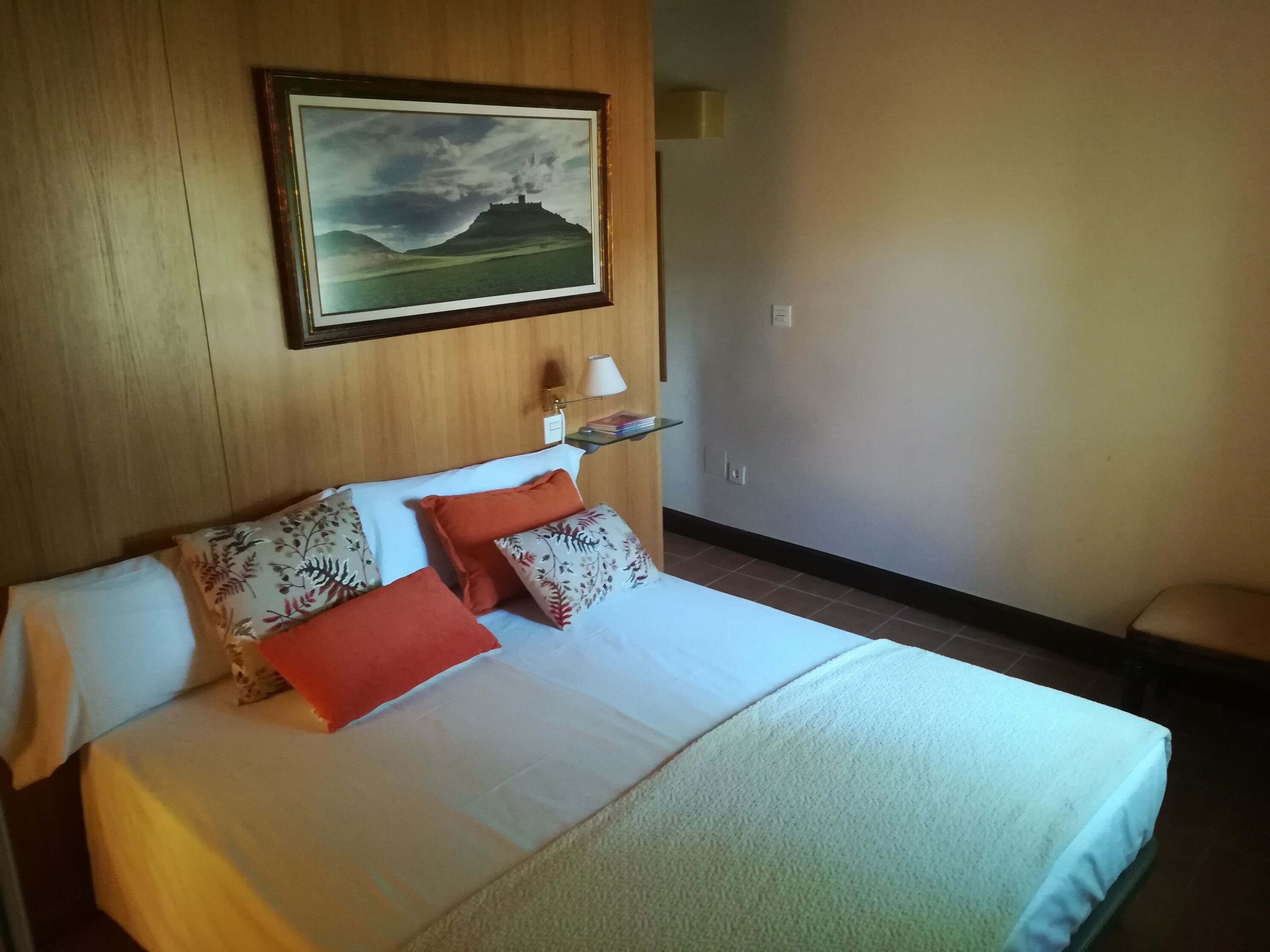 Uma cama bem arrumada com almofadas florais, um quadro de paisagem acima e uma luminária na mesa de cabeceira.