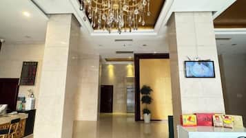 Um saguão de hotel com um lustre, um balcão de recepção, uma tela de televisão e um vaso de planta.