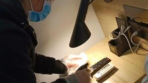 Uma pessoa usando máscara e luvas, trabalhando em uma mesa com uma luminária, um controle remoto e uma caixa de lenços.