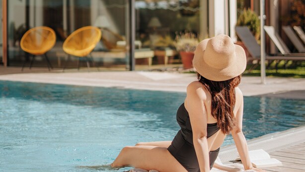 Mulher sentada à beira da piscina, com um edifício moderno ao fundo.