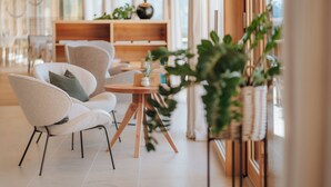 Um interior moderno com área de estar, incluindo cadeiras e uma mesa pequena, um armário de madeira com objetos decorativos, e grandes janelas que permitem entrada de luz natural.