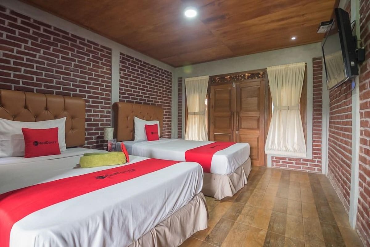 Quarto de hotel com duas camas, cada uma com colcha vermelha e branca, cabeceira de madeira e parede de tijolos.