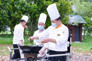 Cozinheiros de uniformes brancos preparam comida ao ar livre com um wok sobre um fogão portátil.