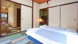 Quarto de hotel com uma cama, um relógio e vista para um corredor com mesa e cadeiras.