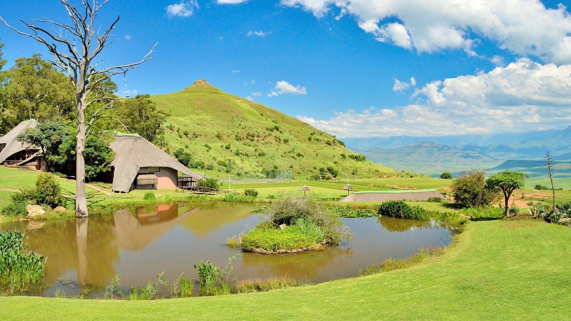 Paisagem rural com um lago, campos gramados e casas com telhados de palha.