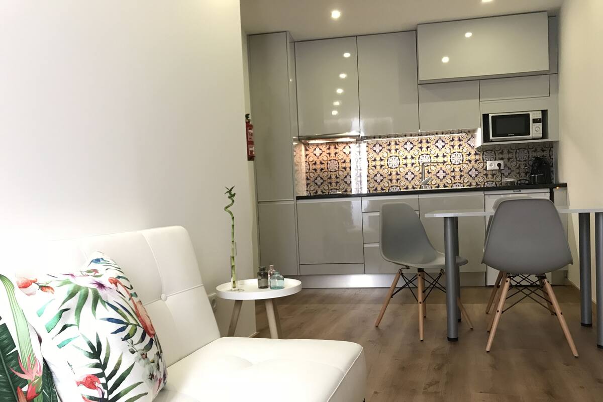 Um espaço de estar compacto com sofá branco, uma pequena mesa redonda e área de cozinha com micro-ondas e painel decorado.