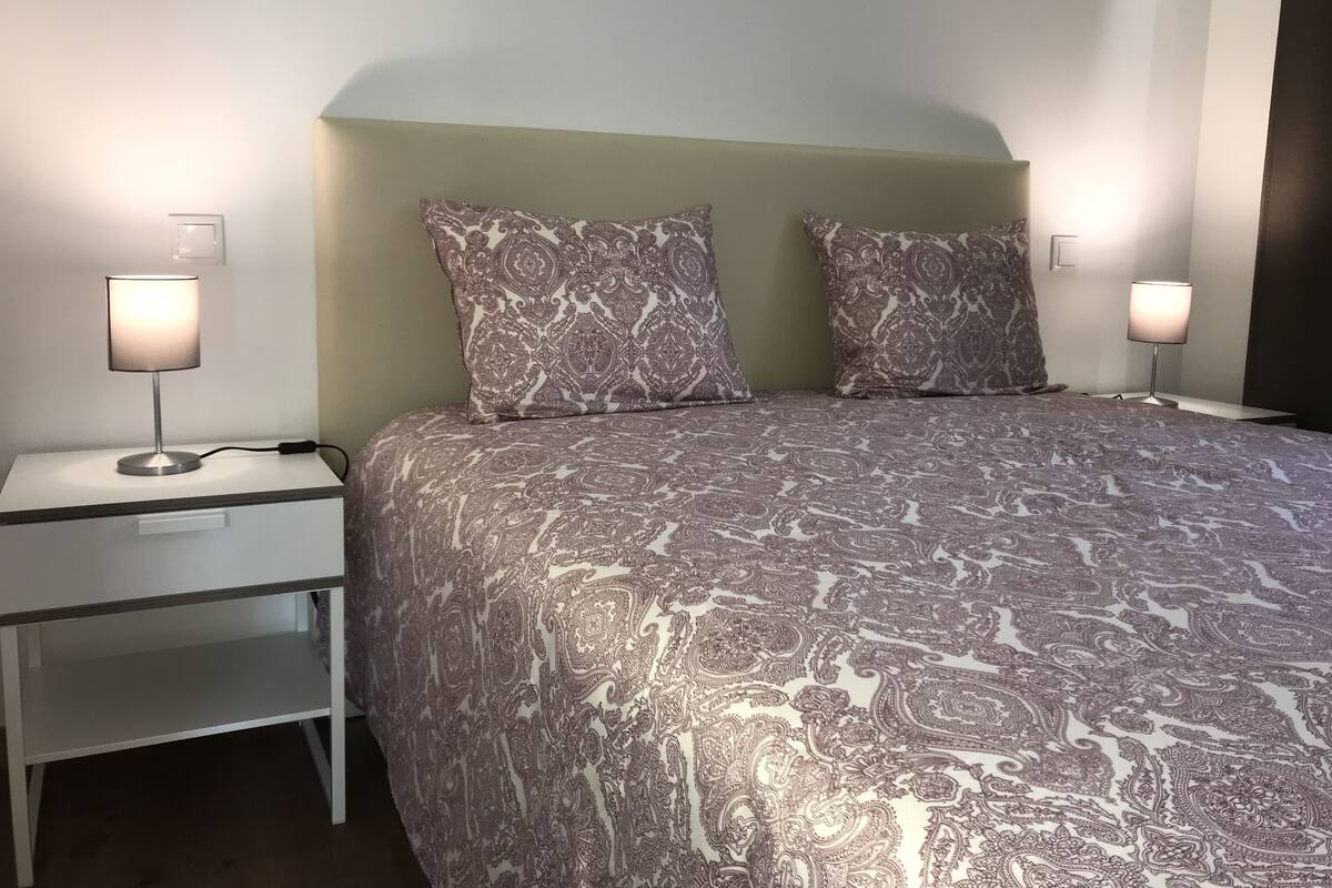 Cama bem arrumada com roupa de cama estampada, duas luminárias de cabeceira e uma pequena mesa de cabeceira branca com uma gaveta.