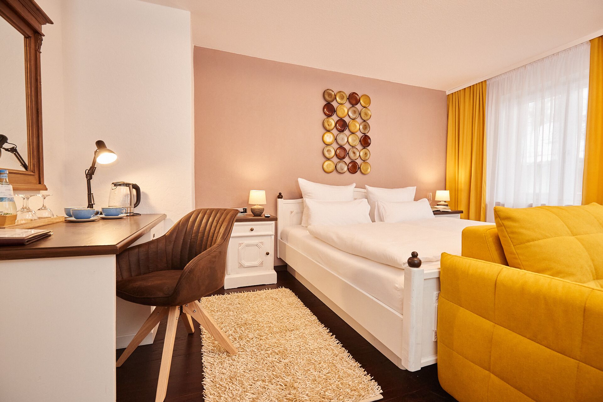 Quarto de hotel com cama, escrivaninha com luminária, cadeira, tapete e uma obra de arte na parede.