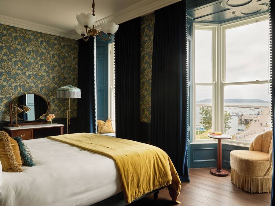 Quarto com cama grande, escrivaninha com luminária, cadeira e vista para o mar pela janela.