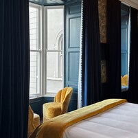 Quarto de hotel com uma cama grande, janela com cortinas, uma cadeira e um espelho.