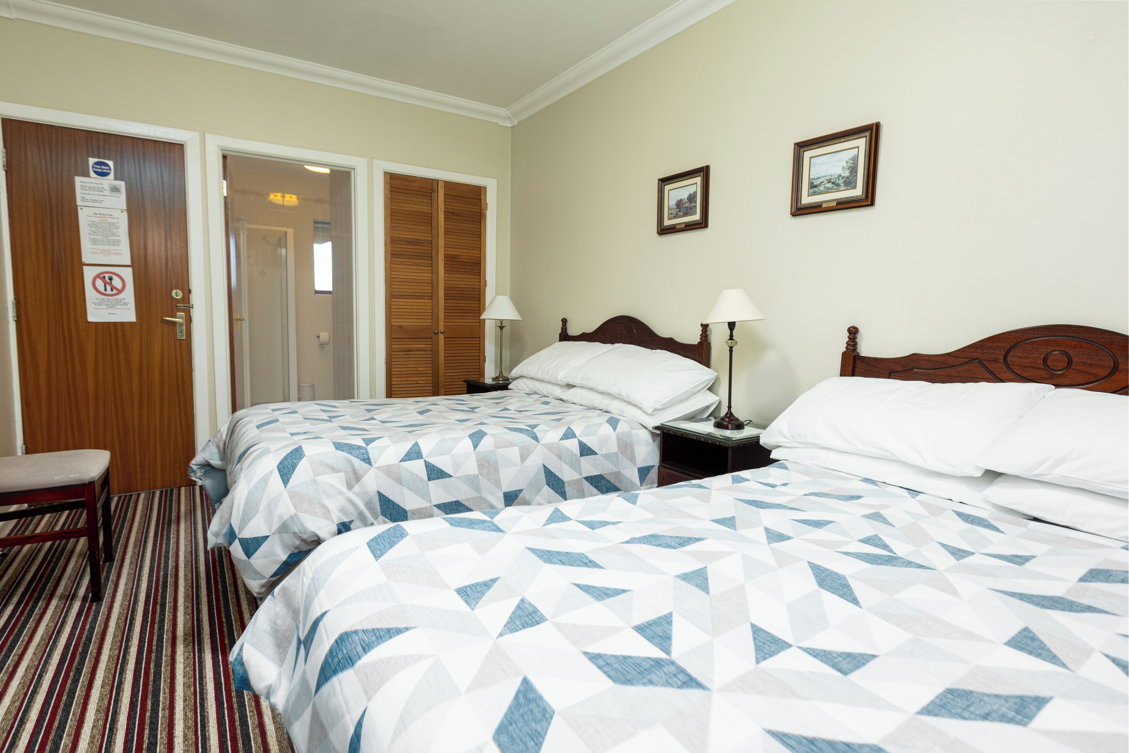 Quarto de hotel com duas camas, um banco, uma porta e duas molduras na parede.