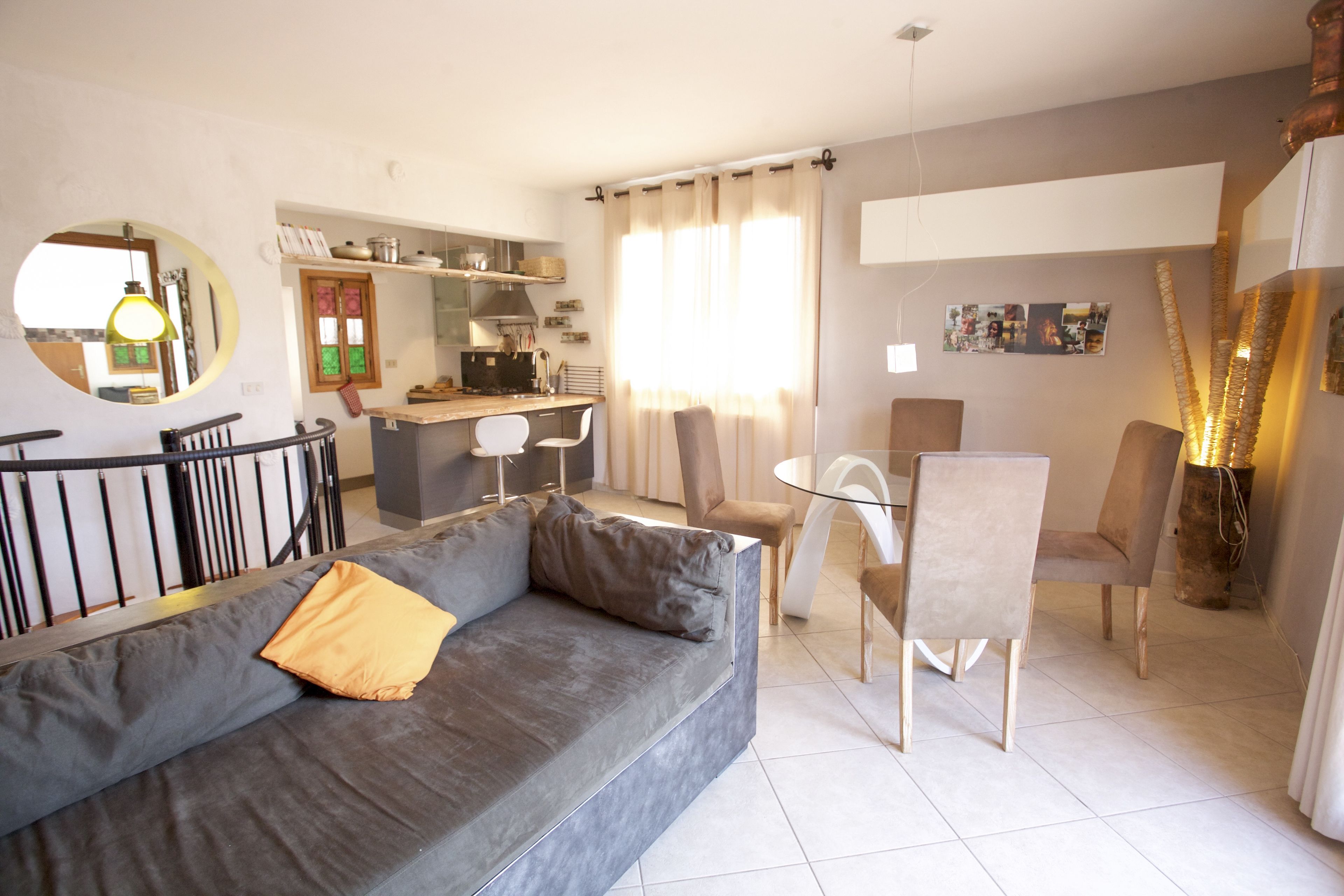Uma sala de estar moderna com um sofá cinzento, uma área de refeições com mesa e cadeiras, e uma cozinha ao fundo.