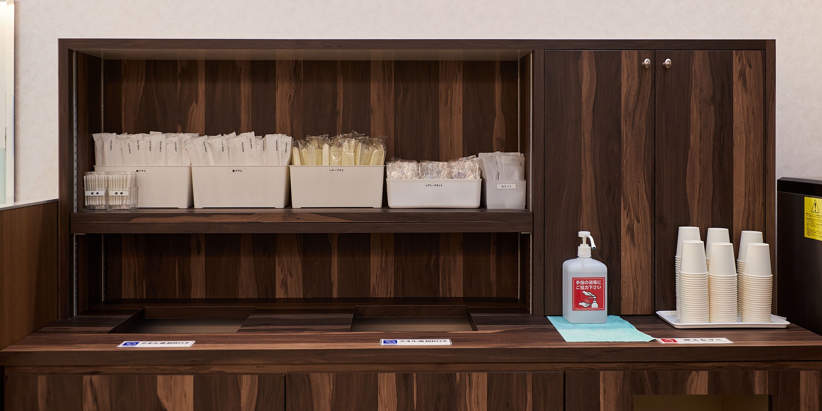Uma área de recepção com painel de madeira ao fundo, balcão com frasco de álcool em gel e pilhas de copos brancos.