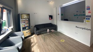 Uma área de recepção com um sofá, uma mesa de centro de vidro, uma máquina de venda automática e uma janela com um letreiro de 'Bem-vindo'.