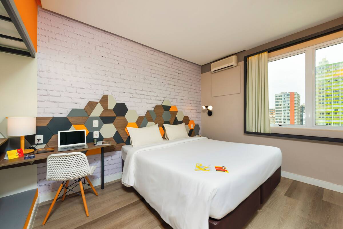 Quarto de hotel com cama, escrivaninha, cadeira e vista para edifícios pela janela.