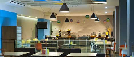 Um restaurante moderno com diversas opções de assentos, incluindo mesas e cabines. Há um balcão com uma variedade de alimentos expostos e uma televisão na parede. O teto apresenta um padrão geométrico, e várias luminárias pendentes o decoram.