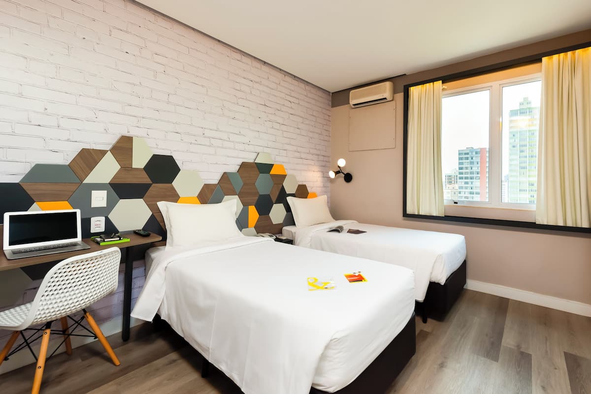 Quarto de hotel com duas camas, uma mesa com um laptop, uma cadeira e vista para edifícios pela janela.