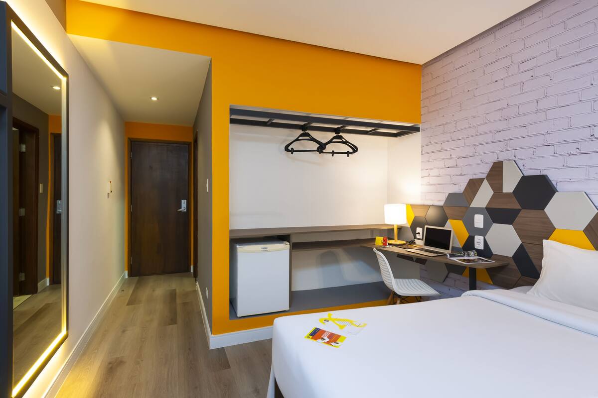 Quarto de hotel com cama, secretária com um laptop, cadeira, mini-frigorífico e cabides fixados na parede.