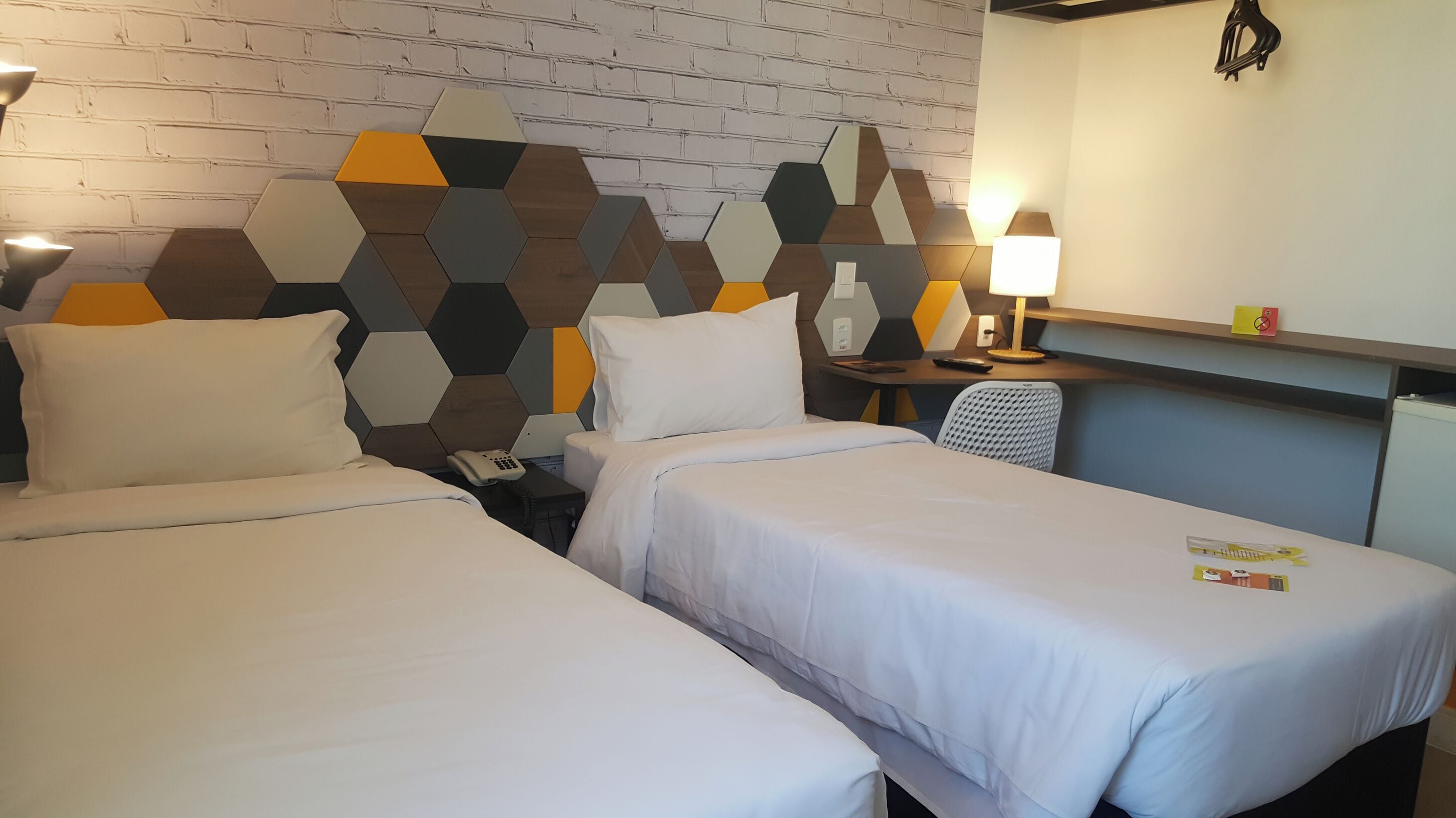 Quarto de hotel com duas camas, cabeceira com padrão de tijolos, escrivaninha com luminária e uma cadeira.