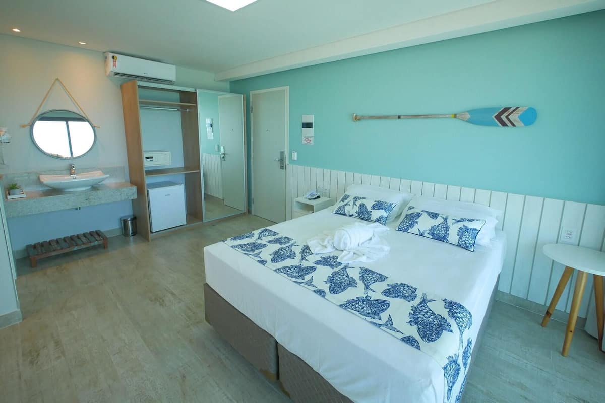 Quarto de hotel moderno com uma cama grande, banheiro com pia e espelho, e uma pequena mesa de cabeceira.