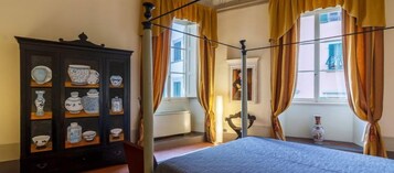 Quarto com uma cama de dossel, um armário de madeira escura com objetos de cerâmica e uma janela com cortinas.