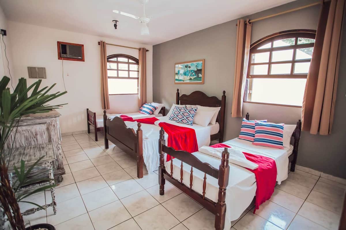 Quarto com duas camas de solteiro, um ventilador de teto, uma janela com cortinas, ar condicionado fixado na parede e um vaso de planta.