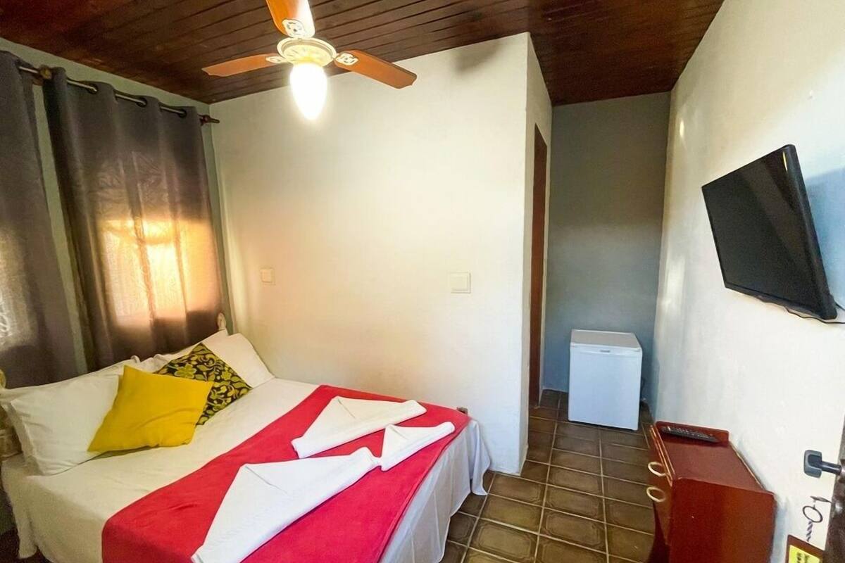 Quarto com cama, ventilador de teto, TV e banheiro ao fundo.