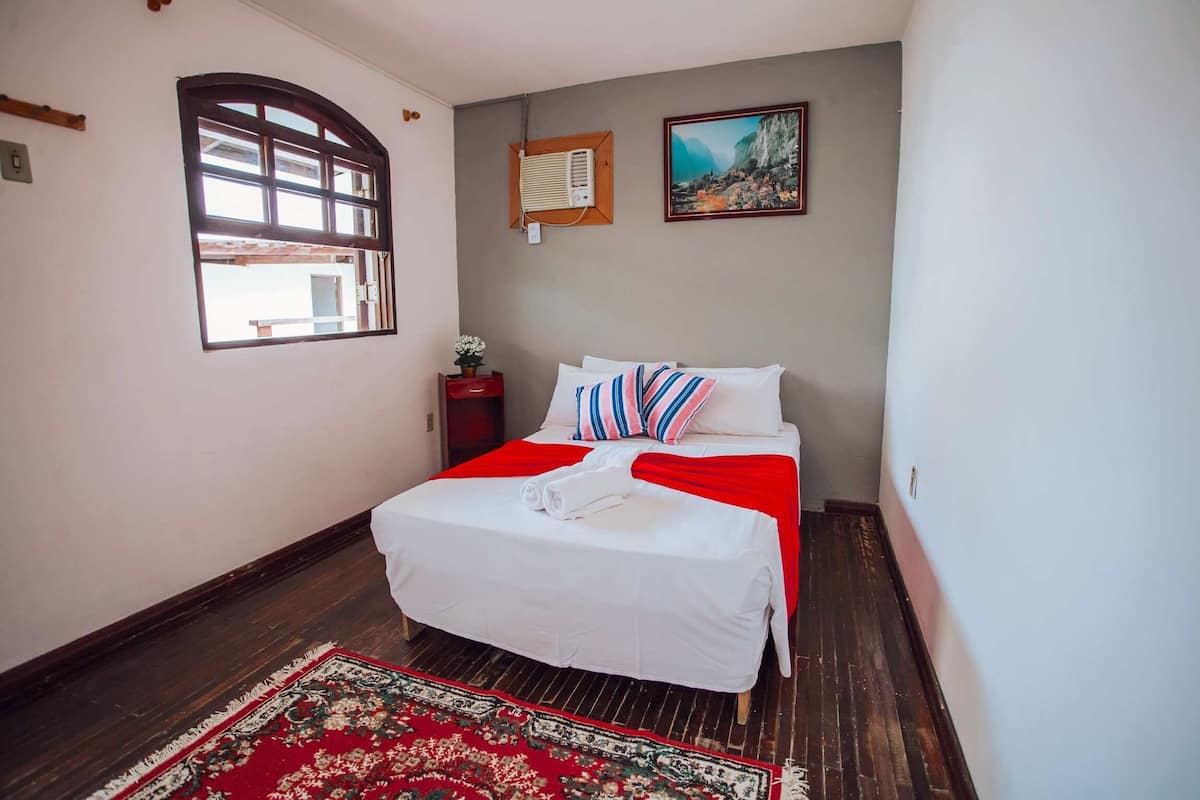 Quarto com cama, janela, ar condicionado fixado na parede e quadro emoldurado.