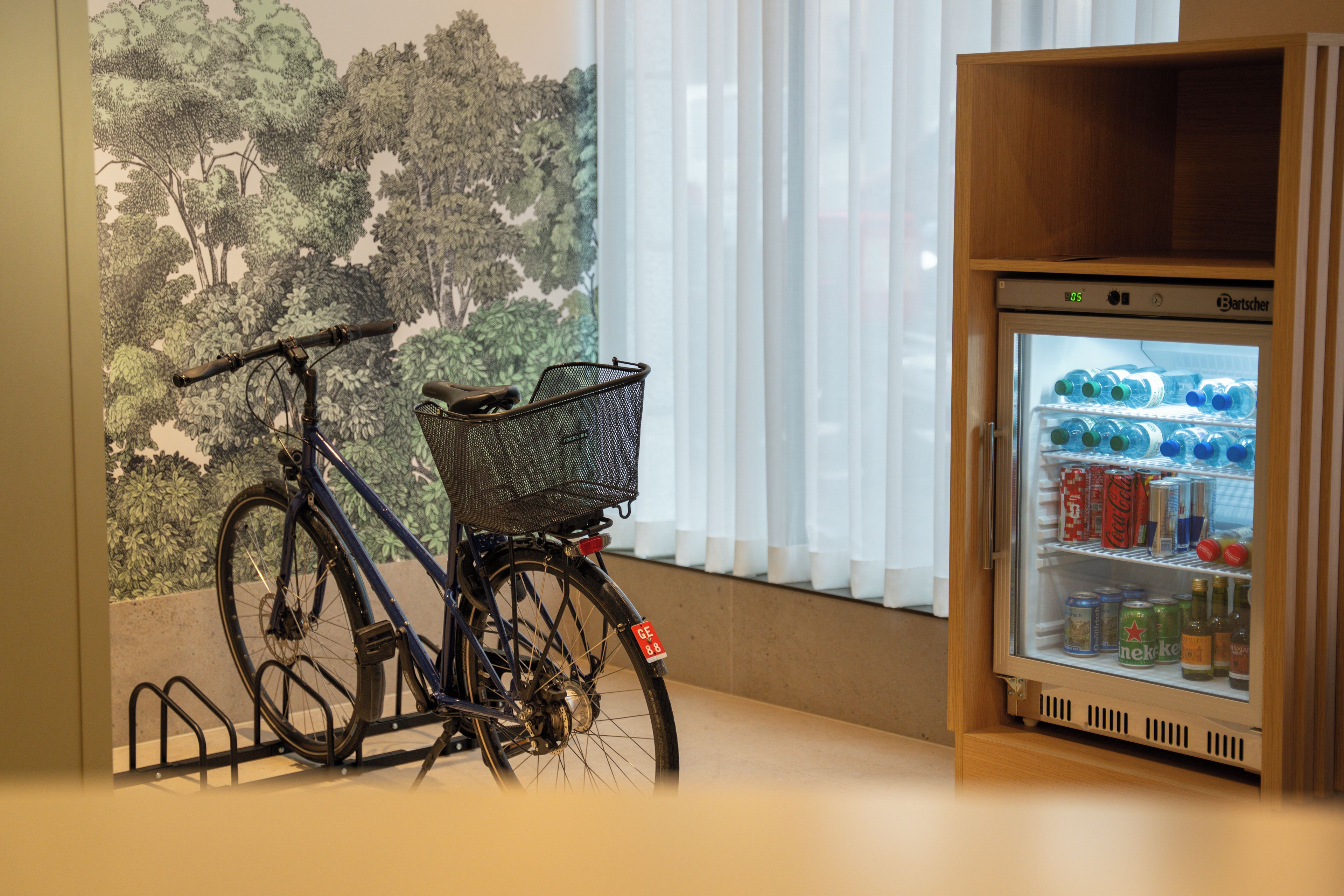 Uma bicicleta com cesto, um muro com mural de árvore e uma geladeira Bartscher abastecida com bebidas.