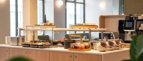 Buffet de café da manhã com diversos alimentos, máquina de café e tela digital.
