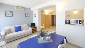 Uma sala de estar com um sofá, uma mesinha com toalha azul, uma planta em vaso e um micro-ondas numa prateleira.