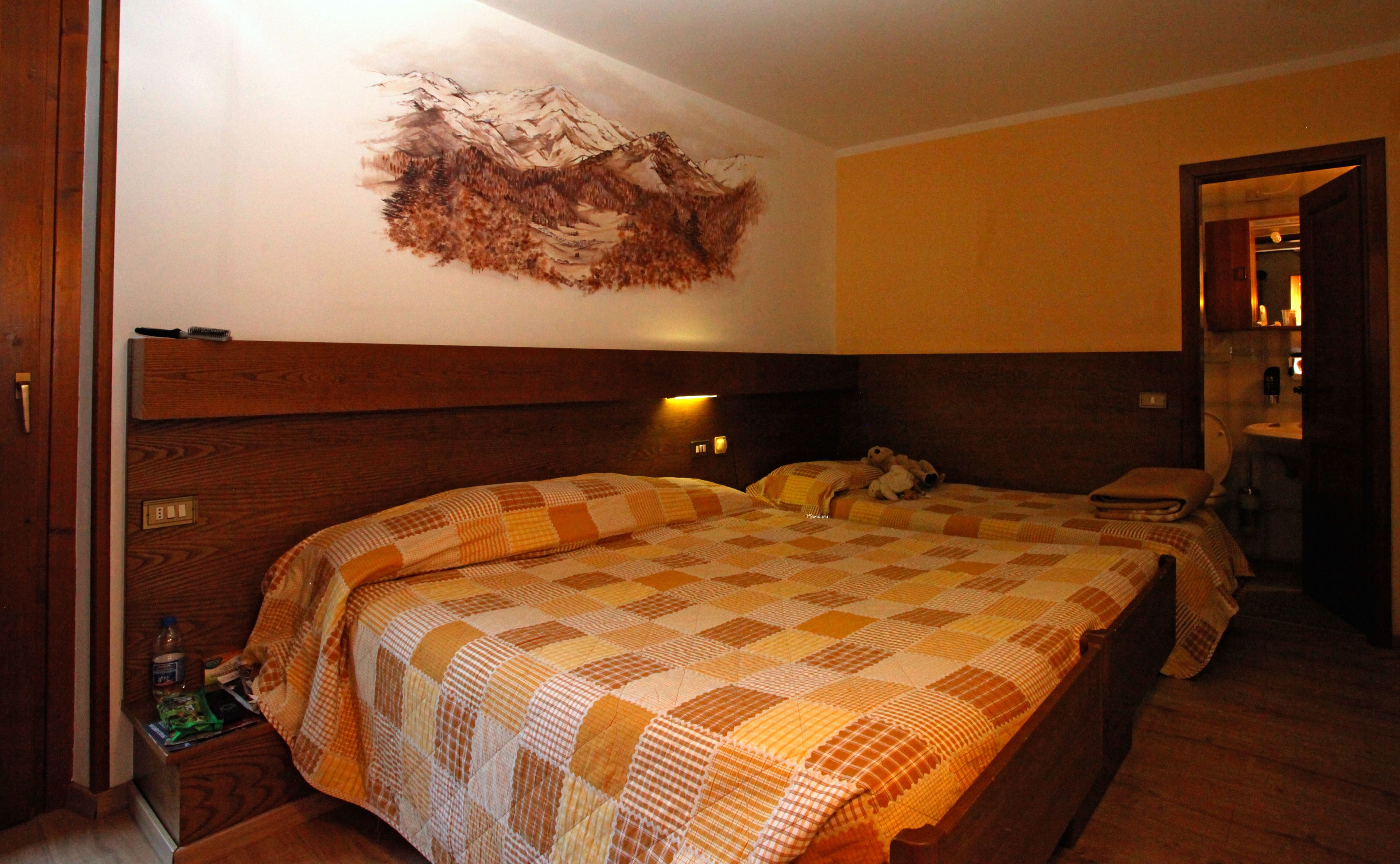 Quarto com uma cama grande, cabeceira de madeira, luminária fixada na parede e banheiro visível através de uma porta aberta.