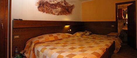 Quarto com uma cama grande, cabeceira de madeira, luminária fixada na parede e banheiro visível através de uma porta aberta.