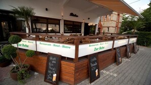 Uma área de estar ao ar livre com mesas e cadeiras de madeira, placas indicando que se trata de um restaurante e um bistrô do hotel, e uma placa com a inscrição 'Zur Rebe' e o preço de 650.