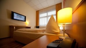 Quarto de hotel com cama, escrivaninha com luminária, televisão na parede e janela com cortinas.