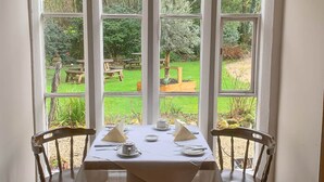 Uma área de jantar com uma mesa posta para quatro pessoas, ladeada por duas cadeiras, e vista para um jardim através de janelas com painéis de vidro decorativos.