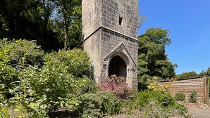 Uma torre de pedra com entrada em arco, rodeada por vegetação exuberante e um caminho de cascalho.